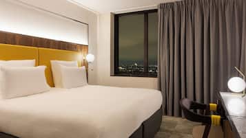 Quarto de hotel com uma cama grande, uma escrivaninha com luminária e vista para a cidade pela janela.