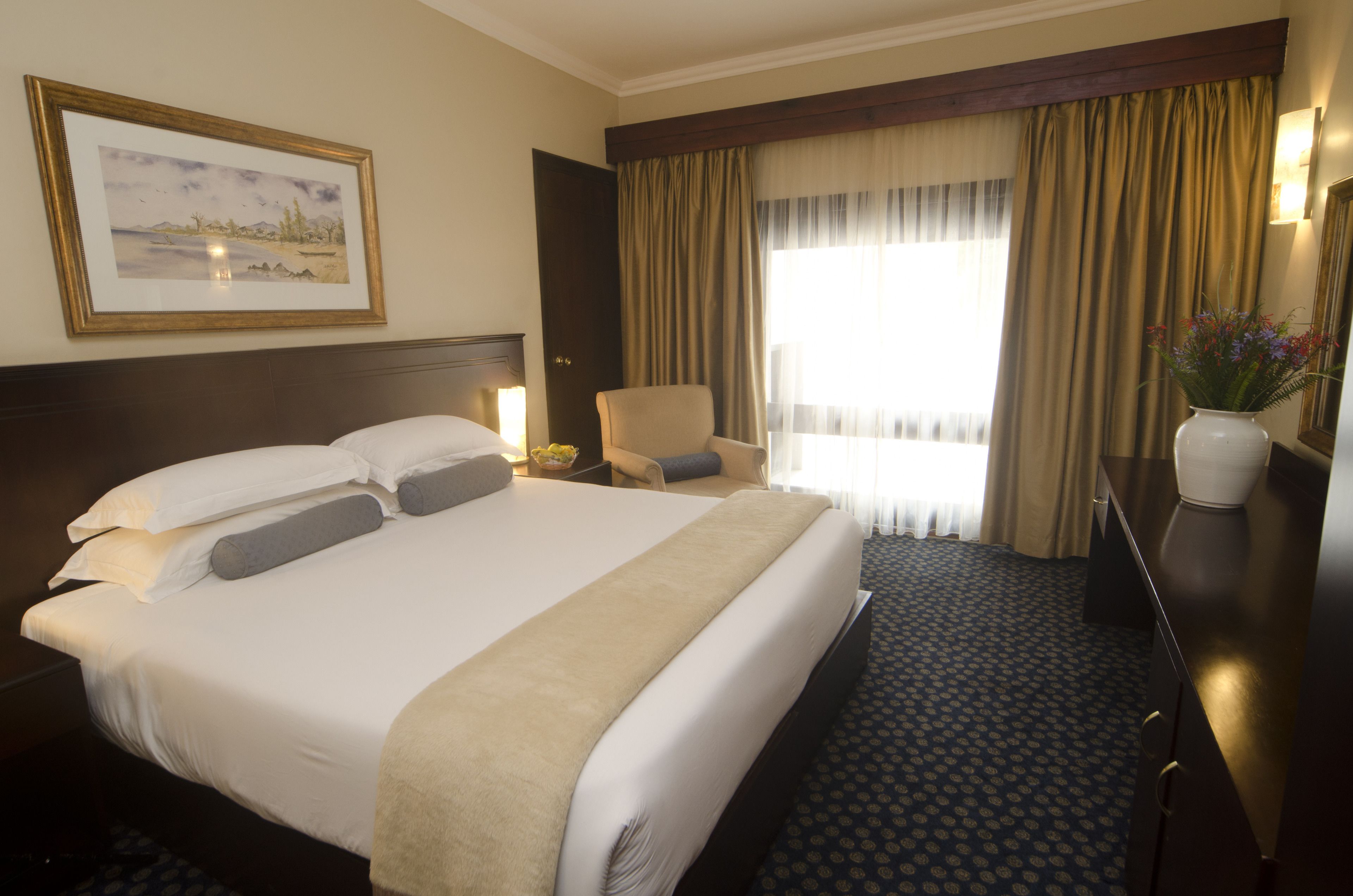 Quarto de hotel com uma cama grande, uma cadeira, uma escrivaninha e uma janela com cortinas.