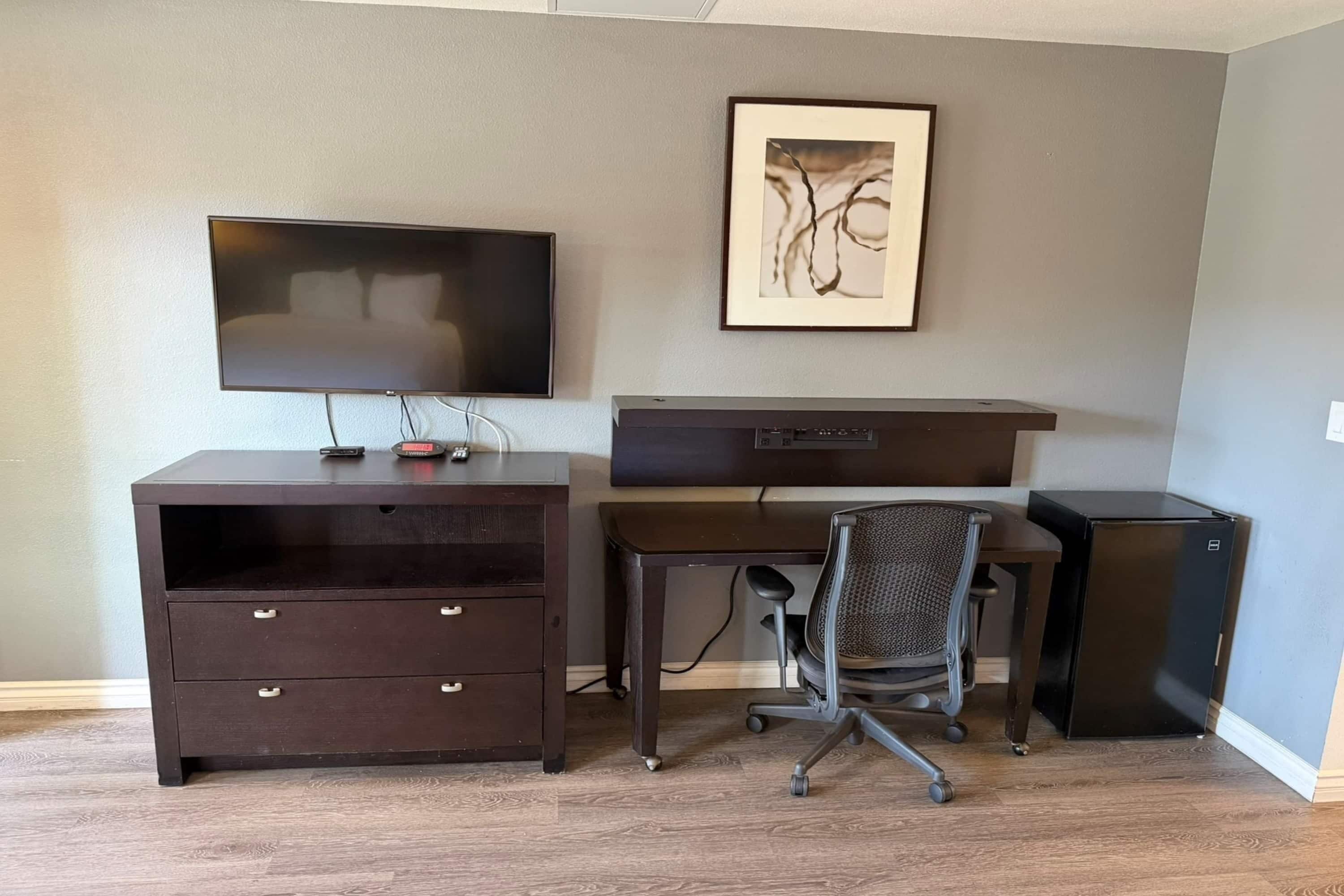 Um espaço de escritório moderno com uma mesa de madeira escura, uma cadeira de escritório preta, uma TV de tela plana, um pequeno refrigerador preto e uma obra de arte abstrata emoldurada na parede.