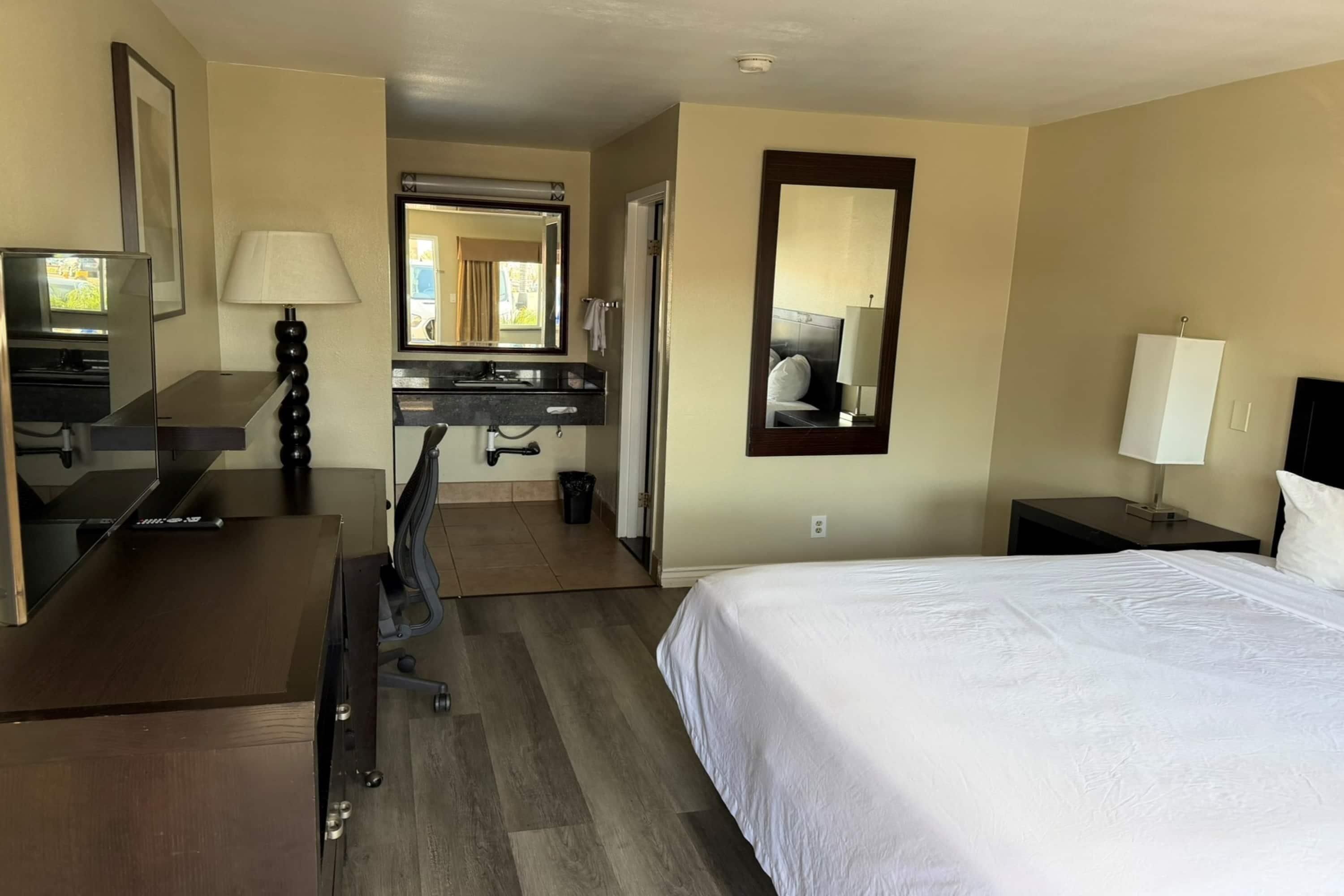 Quarto de hotel com uma cama, uma escrivaninha, uma cadeira, um espelho, uma mesa de cabeceira e um abajur.