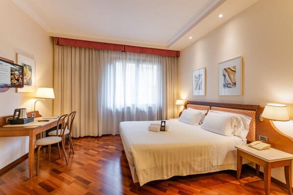 Quarto de hotel com uma cama grande, uma escrivaninha com cadeira, uma luminária e uma janela com cortinas.