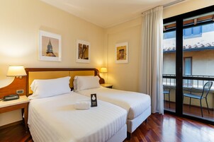Quarto de hotel com duas camas, uma mesa de cabeceira com telefone, abajur e quadros na parede.