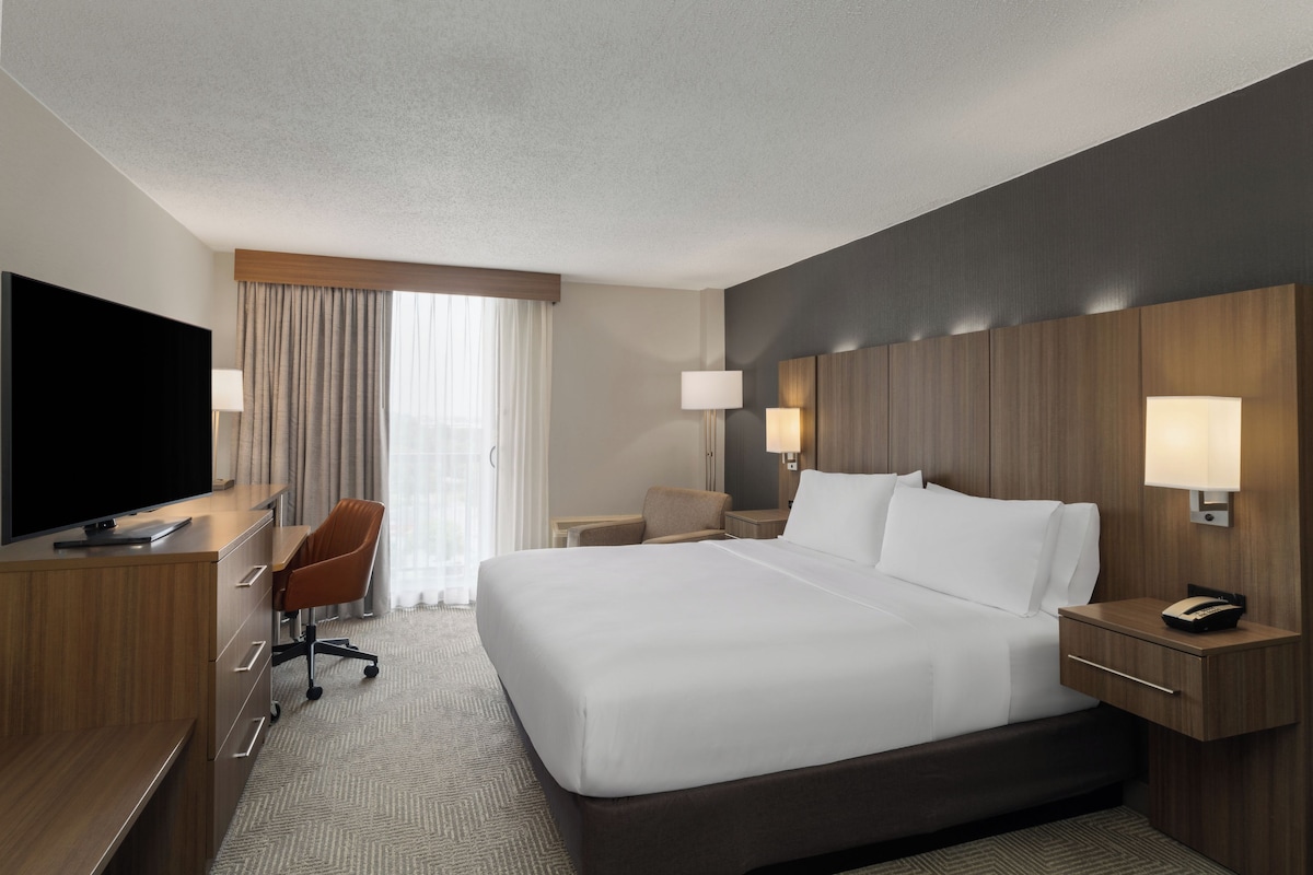 Quarto de hotel com cama grande, escrivaninha com TV de tela plana, uma cadeira, duas luminárias de cabeceira e mesa de cabeceira com telefone.