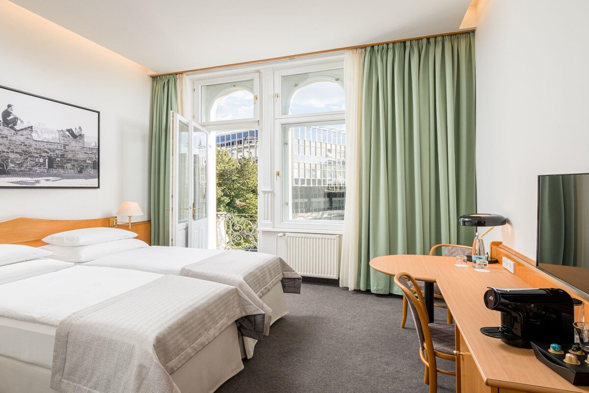 Quarto de hotel com duas camas, uma mesa de jantar, cafeteira e uma janela grande com cortinas.