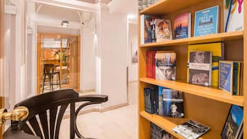 Interior de biblioteca com estantes, uma cadeira preta e uma mesa com abajur.