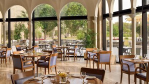 Uma sala de jantar ampla com várias mesas e cadeiras, grandes janelas com vista para o exterior e um interior bem iluminado.