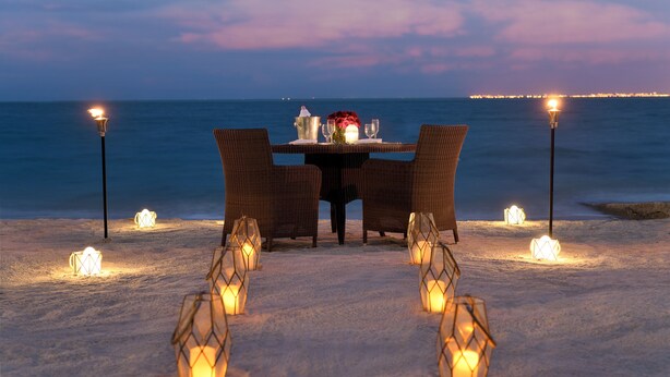 Mesa de jantar posta para dois, com cadeiras de vime, um arranjo de velas e vista para o oceano ao entardecer.