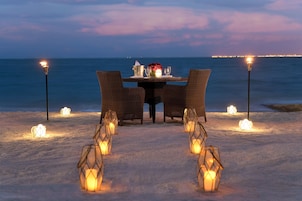 Mesa de jantar posta para dois, com cadeiras de vime, um arranjo de velas e vista para o oceano ao entardecer.