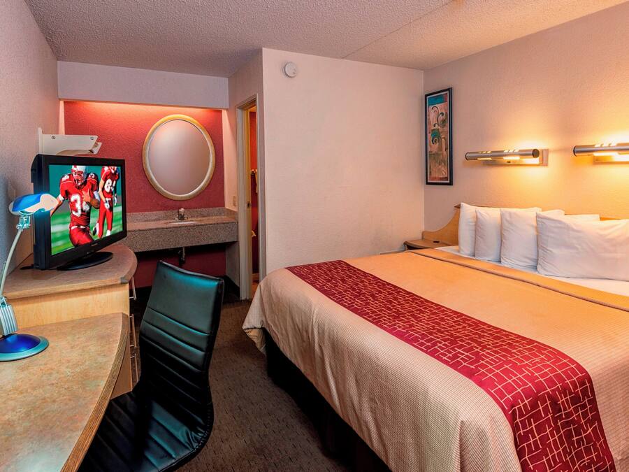 Quarto de hotel com cama, escrivaninha com televisão, cadeira, espelho e luminária.
