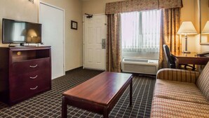 Quarto de hotel com televisão em suporte de madeira, sofá, mesa pequena, escrivaninha com luminária e janela com cortinas.