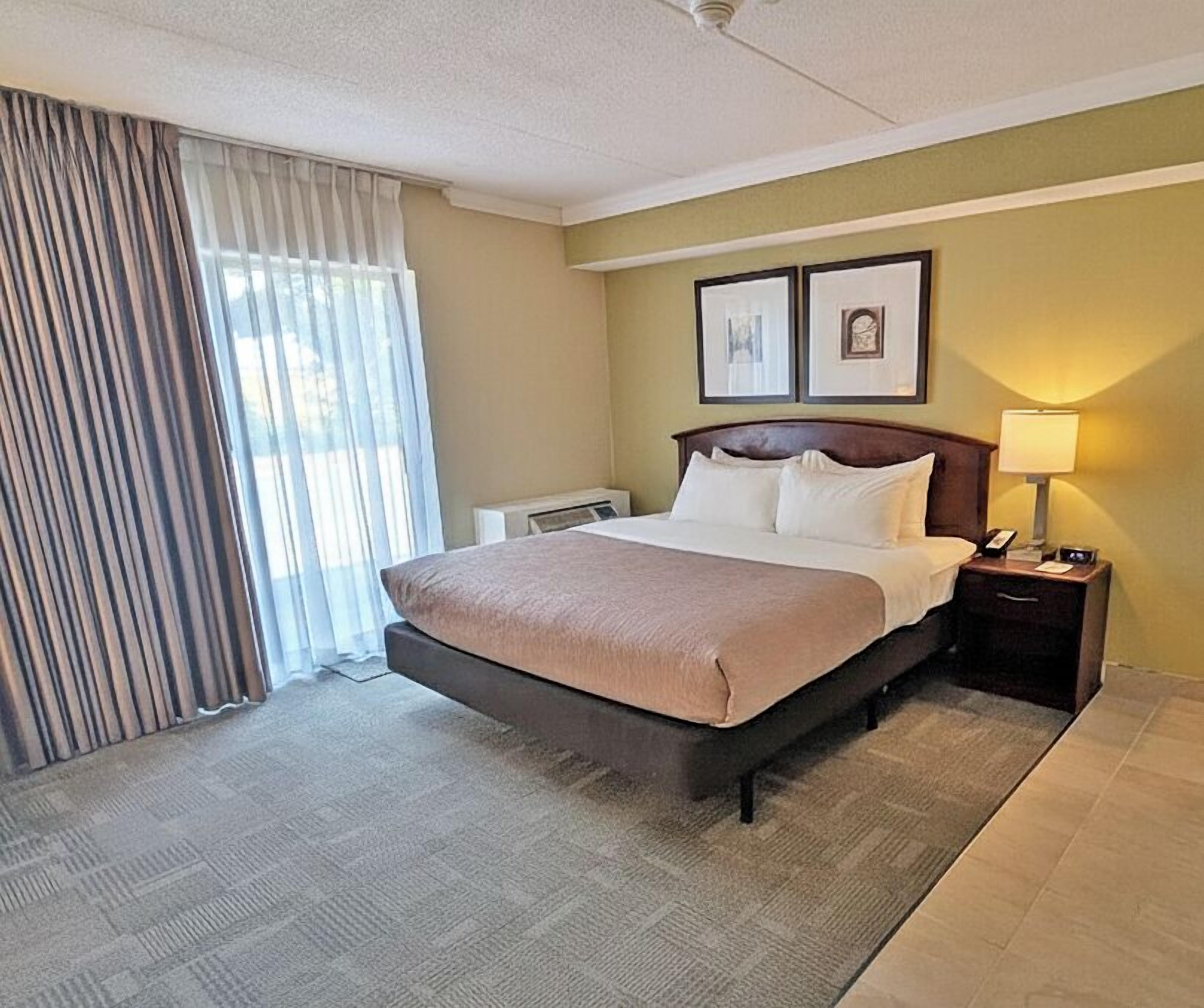 Quarto de hotel com uma cama grande, uma mesa de cabeceira com abajur e uma janela com cortinas.