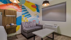 Quarto de hotel moderno com sofá cinza, uma mesa pequena, mesa de cabeceira de madeira com telefone, painel mural abstrato colorido e uma tela grande e vazia na parede.