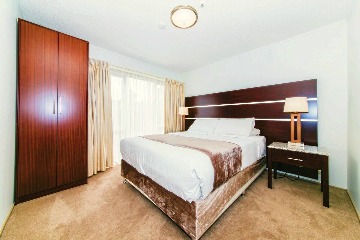 Quarto de hotel com uma cama grande, um guarda-roupa de madeira, uma mesinha de cabeceira com luminária e cabeceira com iluminação embutida.