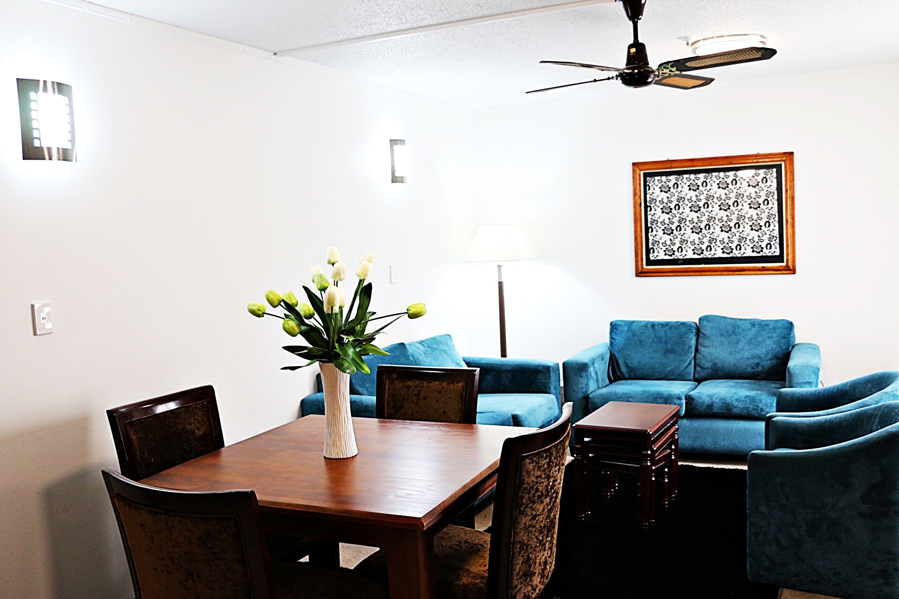 Uma sala de jantar com uma mesa e cadeiras de madeira, um vaso com flores, um sofá azul e um quadro na parede.