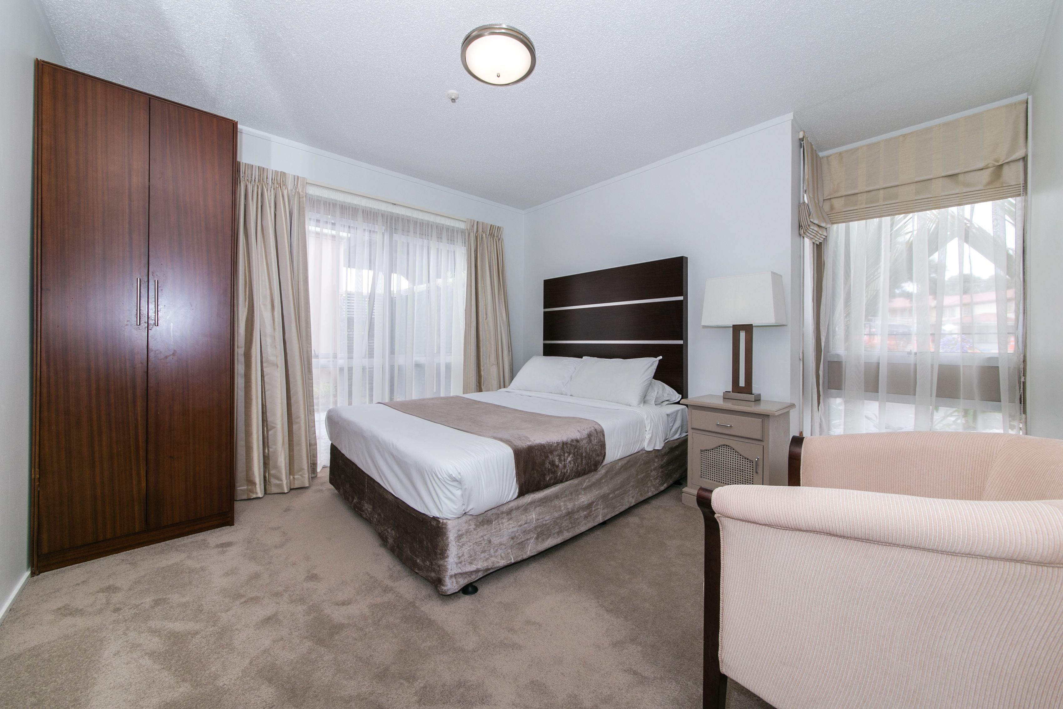 Quarto de hotel com cama, guarda-roupa, mesa de cabeceira, luminária e janela com cortinas.