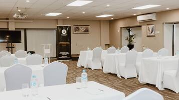 Sala de conferência com mesas e cadeiras brancas, garrafas de água e tela de projeção.