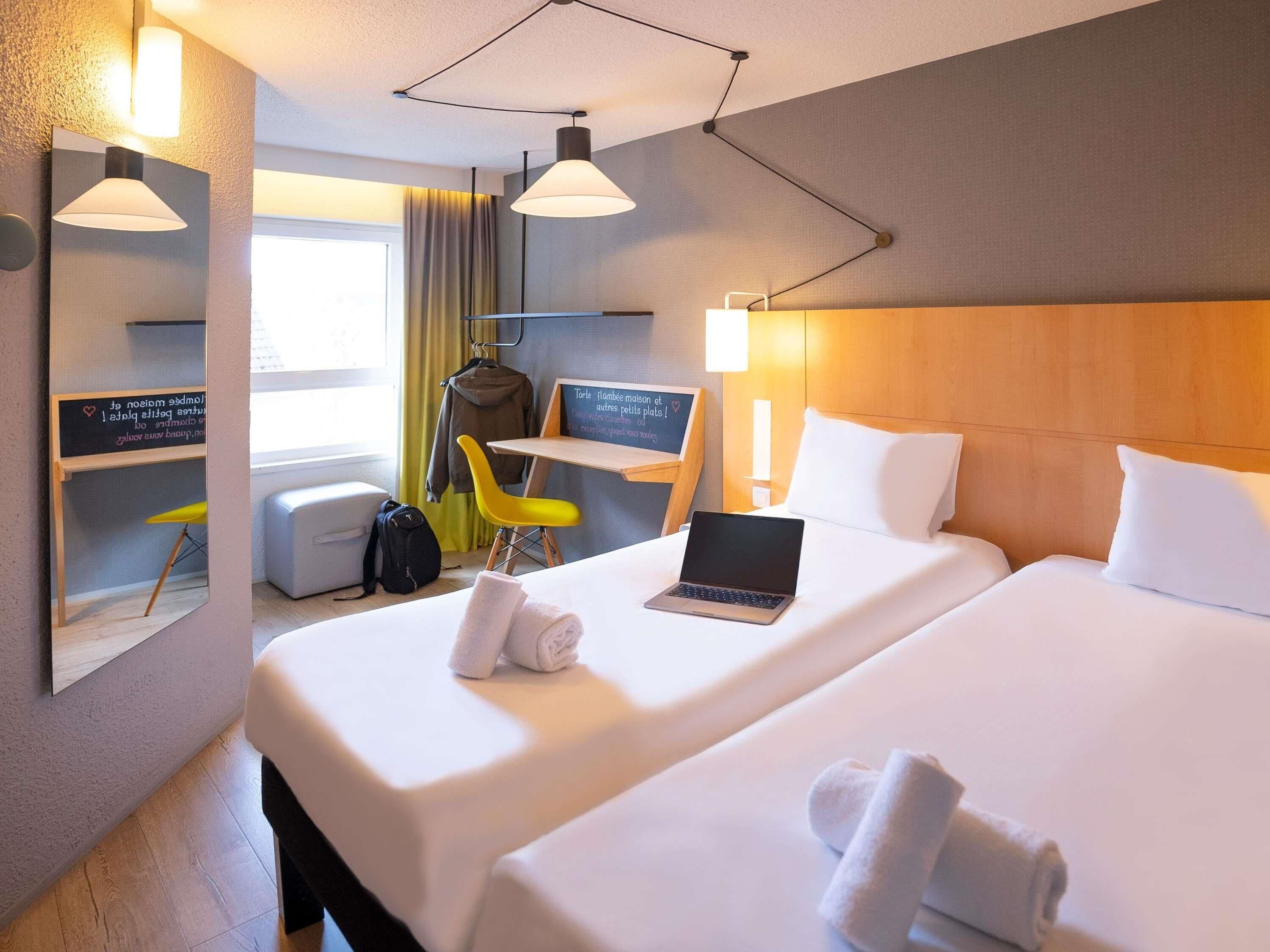 Quarto de hotel com cama, escrivaninha com um laptop, cadeira, cabideiro e espelho.