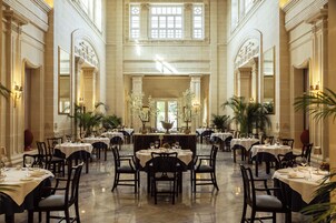 Uma ampla sala de jantar com várias mesas e cadeiras, grandes janelas e detalhes arquitetônicos clássicos.