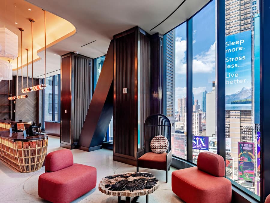Um hall de hotel moderno com teto curvo, balcão de recepção, assentos vermelhos e grandes janelas com vista para a cidade.