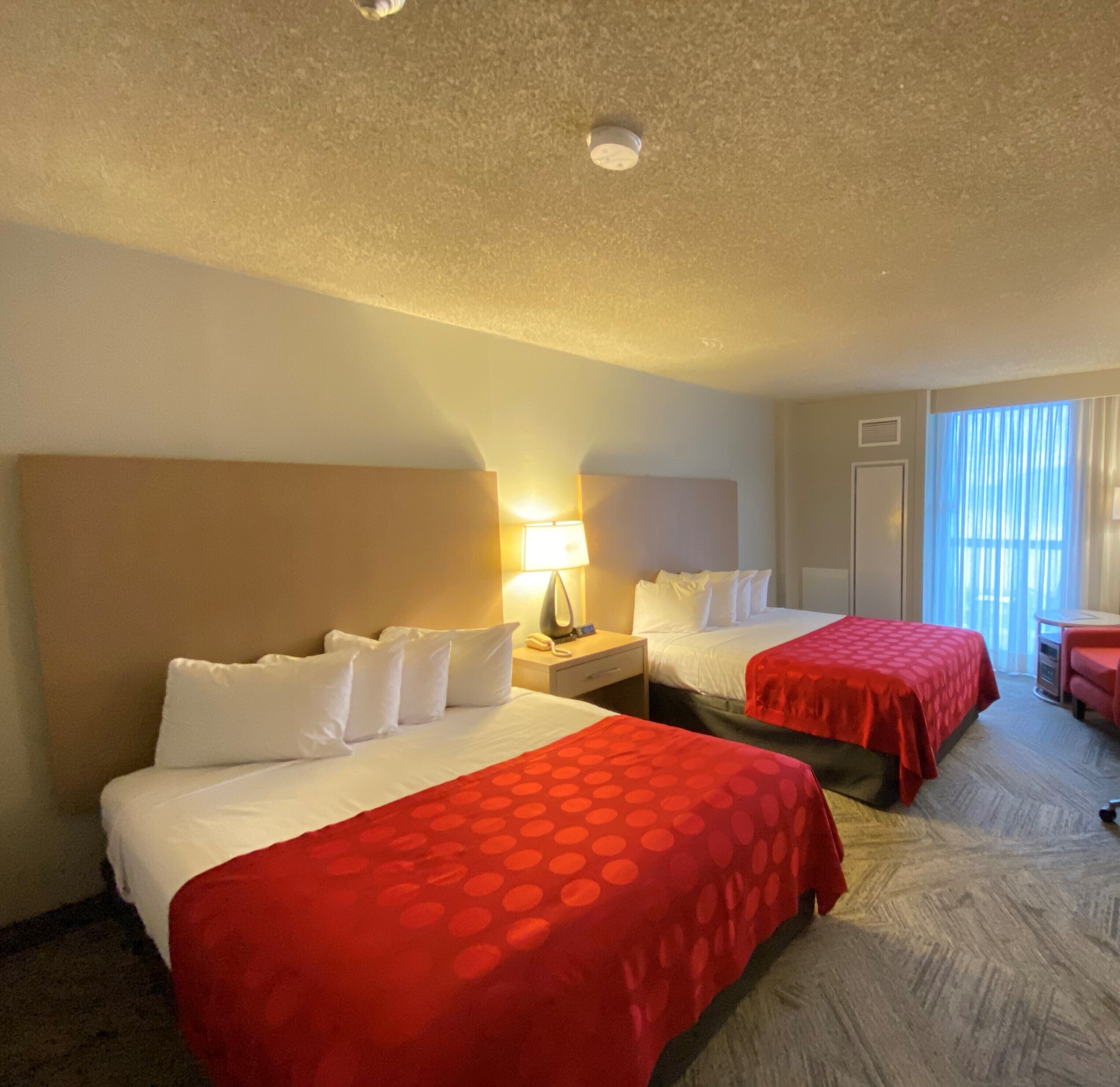 Quarto de hotel com duas camas, uma escrivaninha, uma cadeira e uma janela com cortinas.
