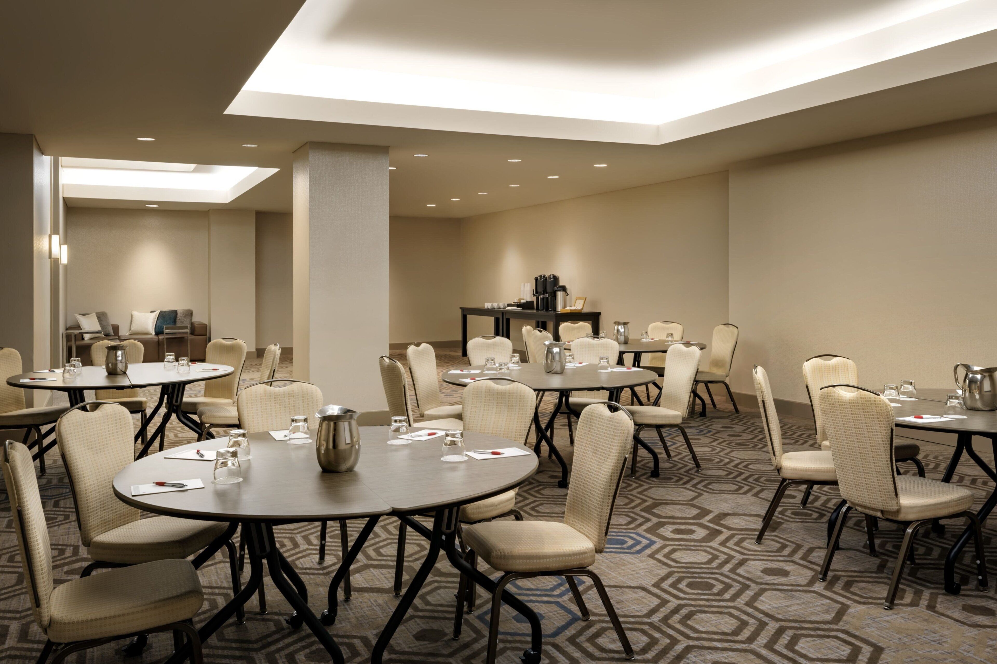Sala de conferências com várias mesas e cadeiras, equipada com máquinas de café e copos de água.