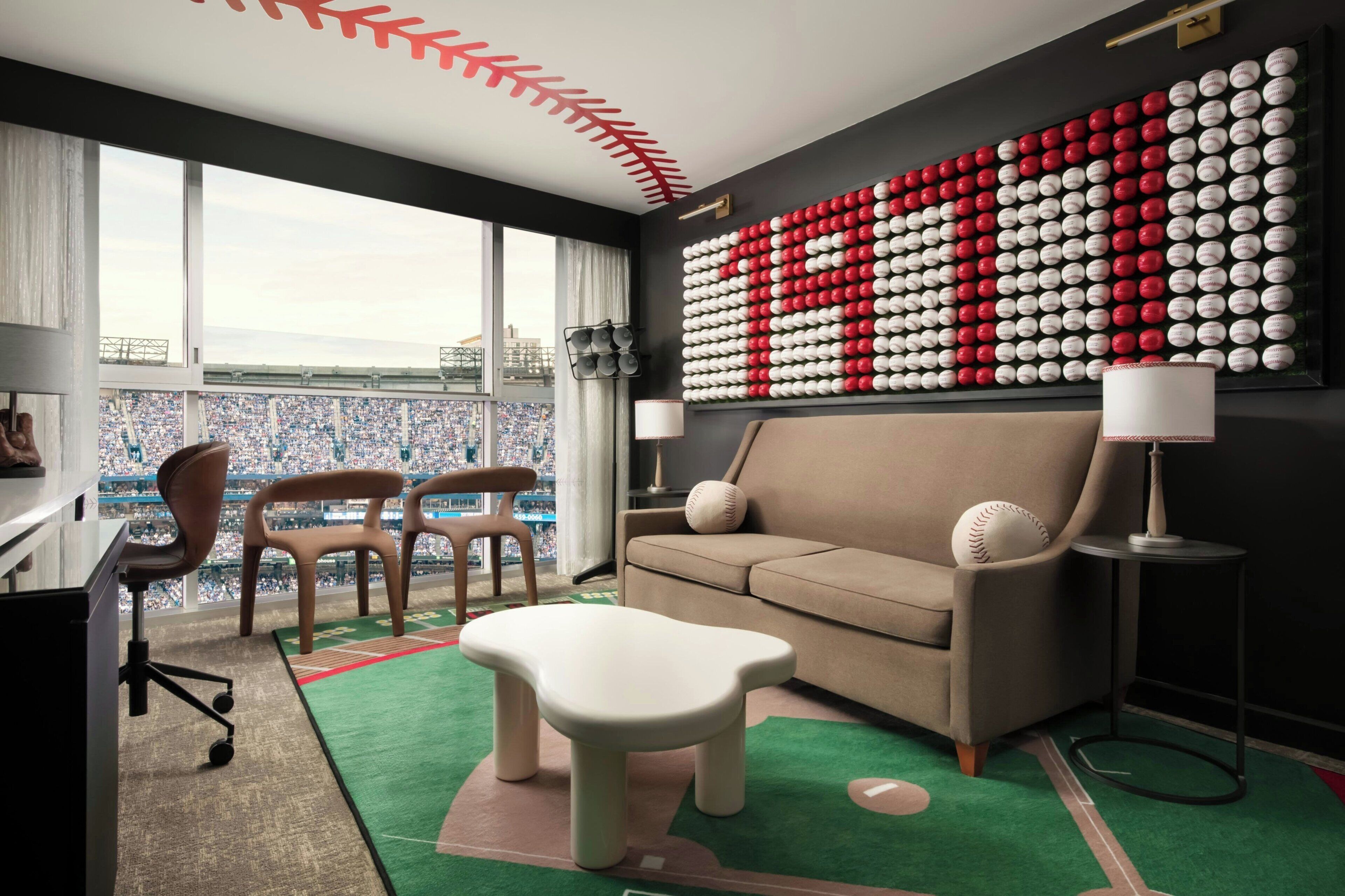Uma sala de estar moderna com uma janela grande, uma obra de arte com tema de beisebol, um sofá, uma mesa de centro e uma escrivaninha com uma cadeira.