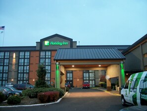 Um hotel Holiday Inn com uma entrada ampla, estacionamento e uma bandeira ao fundo.