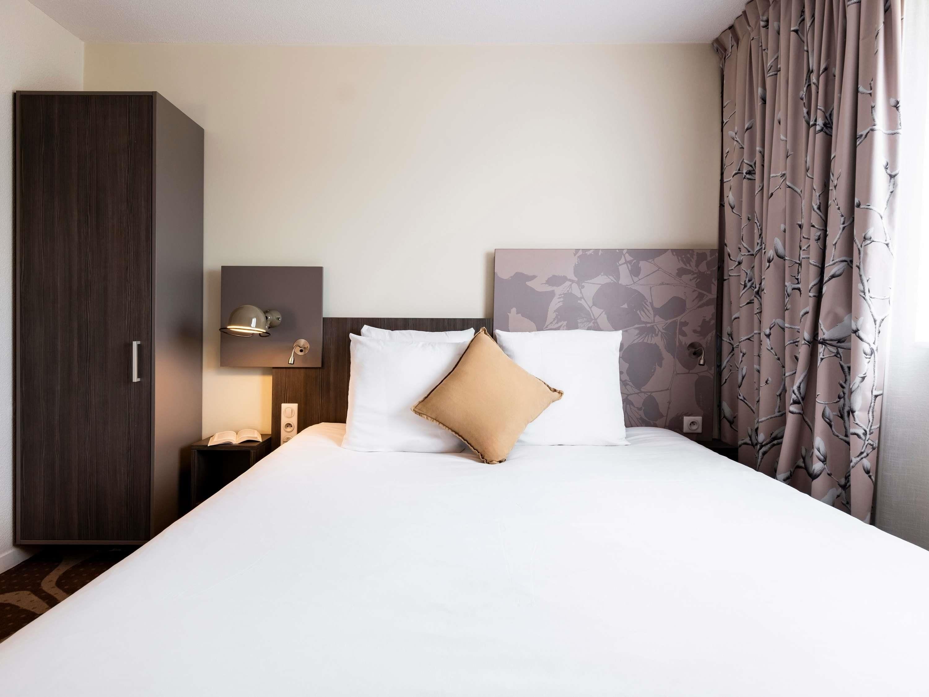 Quarto de hotel com cama, guarda-roupa, mesa de cabeceira e uma luminária fixada na parede.