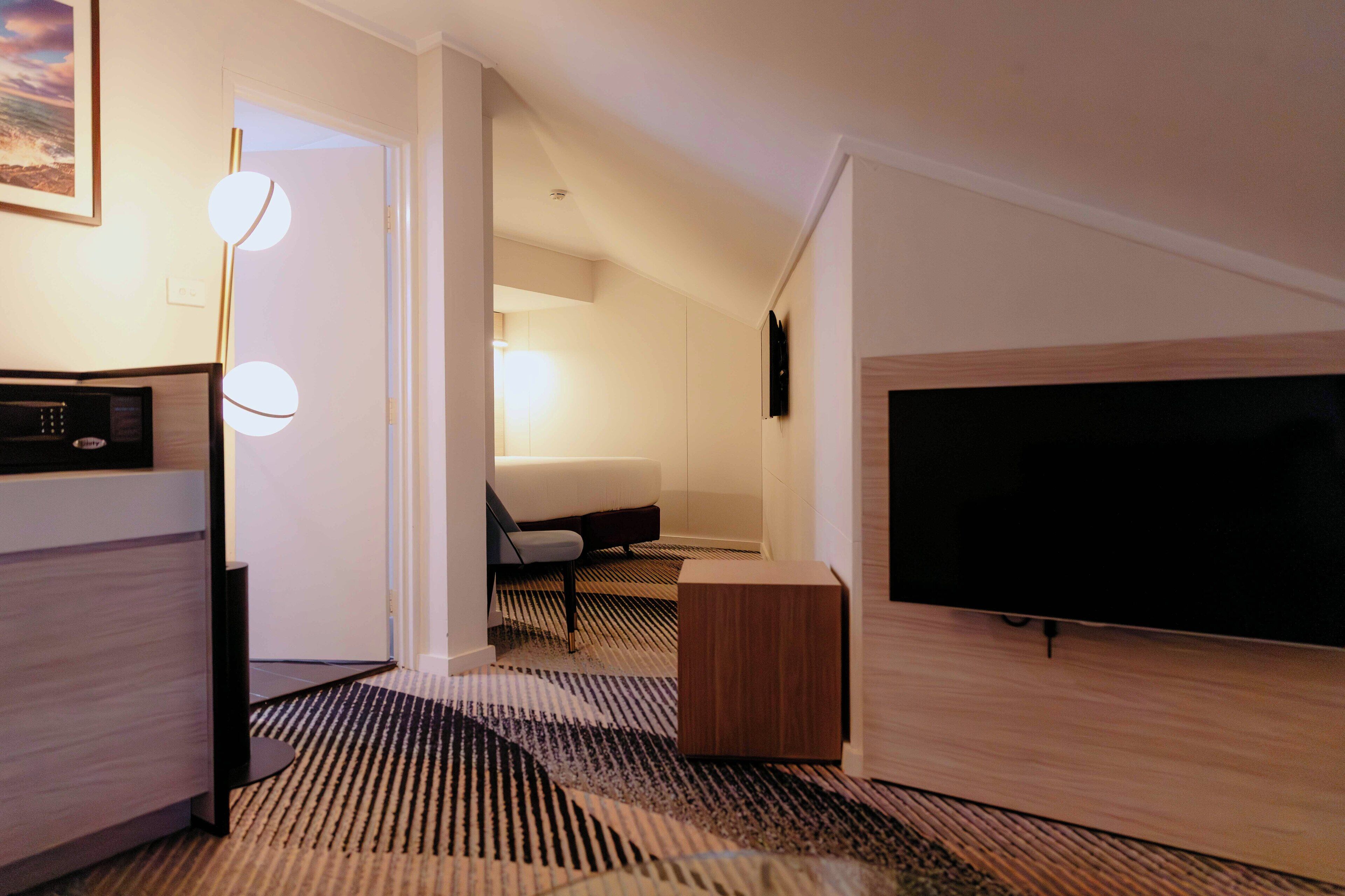 Quarto de hotel moderno com uma televisão de tela plana grande, um sofá, uma pequena mesa lateral de madeira e um tapete estampado.