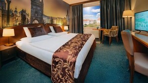 Quarto de hotel com uma cama grande, duas mesas de cabeceira com abajures, uma escrivaninha com cadeira e uma janela ampla com vista para edifícios.