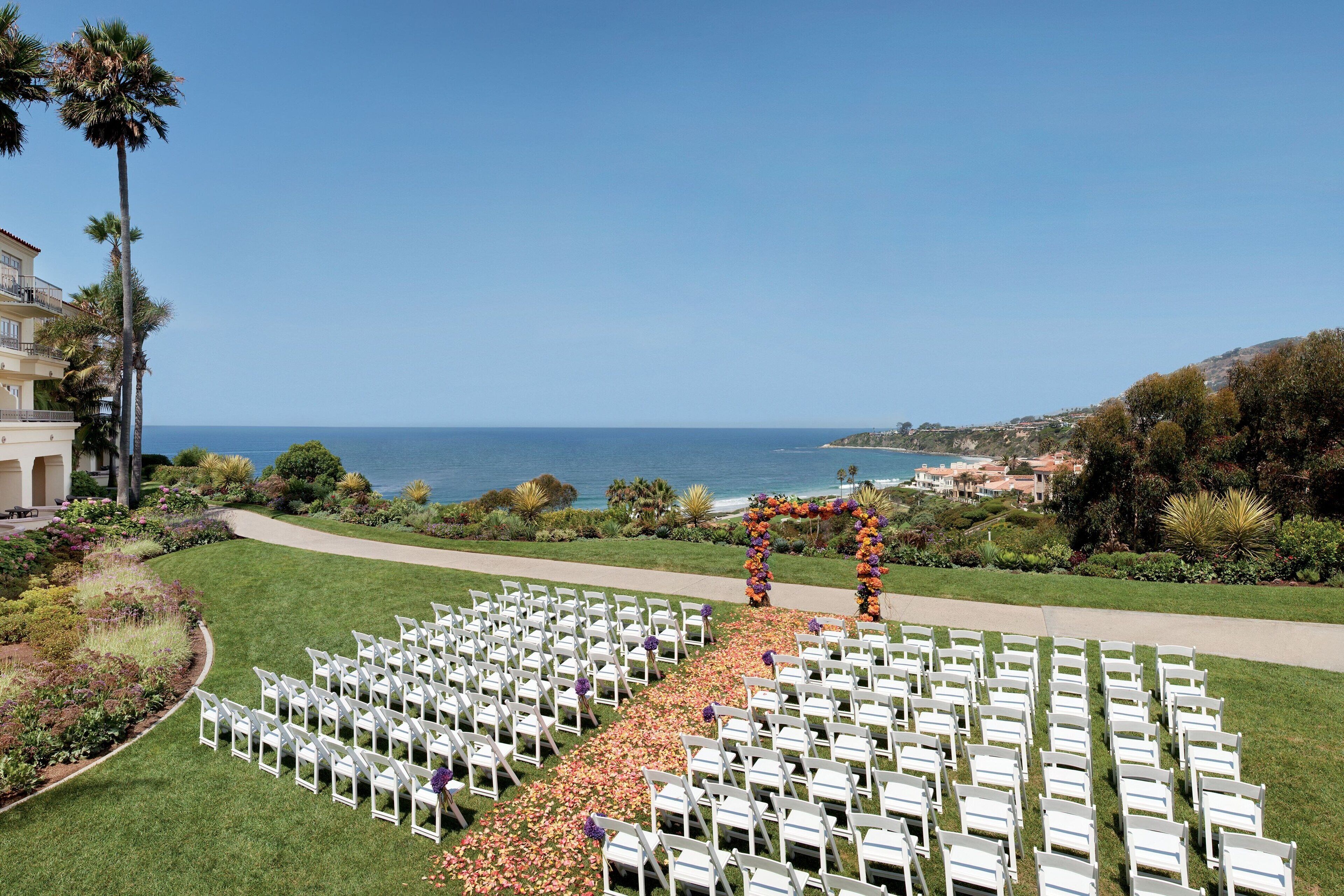 Preparação para uma cerimônia de casamento com fileiras de cadeiras brancas e um arco floral, com vista para uma paisagem costeira e edifícios ao longe.
