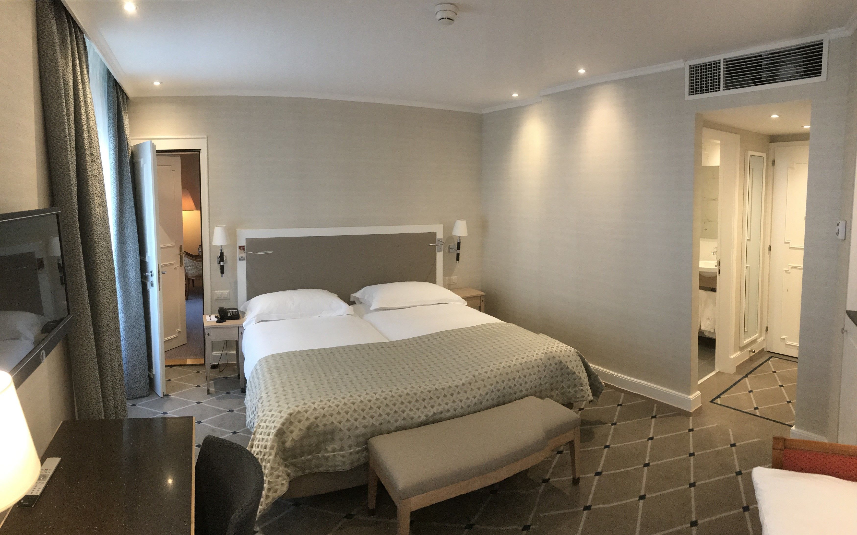 Quarto de hotel com cama, mesa de cabeceira, luminária e um banco.