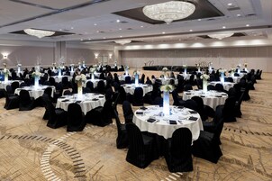 Uma sala de banquetes com mesas redondas, cadeiras pretas e centros de mesa florais.