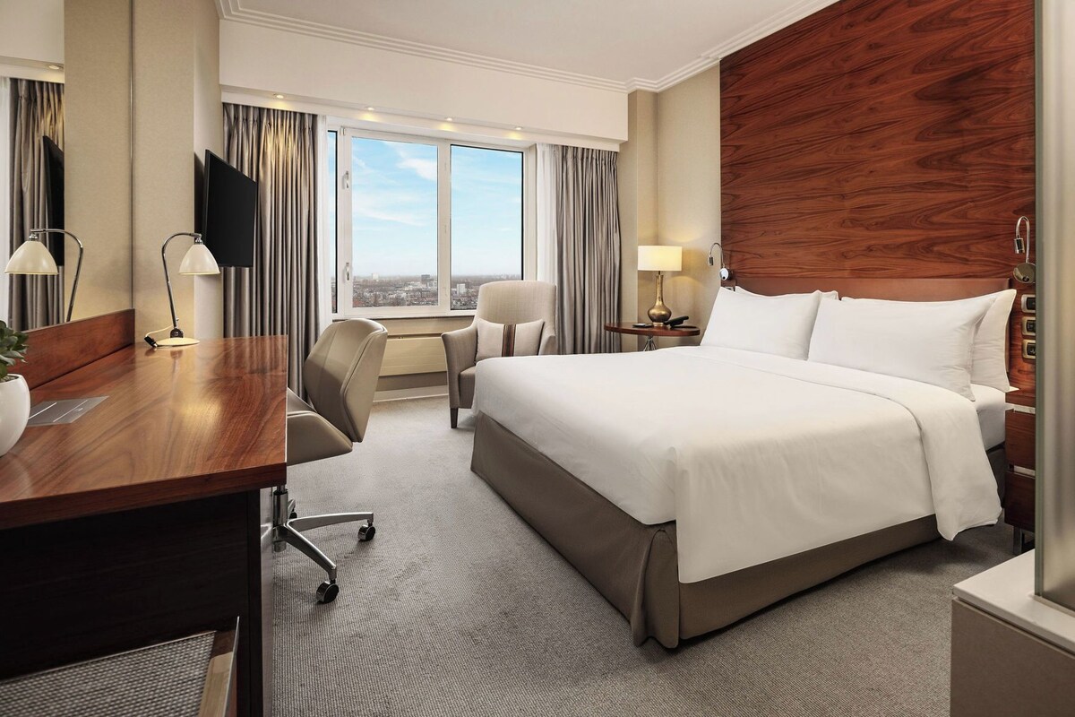 Quarto de hotel com uma cama grande, uma escrivaninha com cadeira, televisão e vista para a cidade.