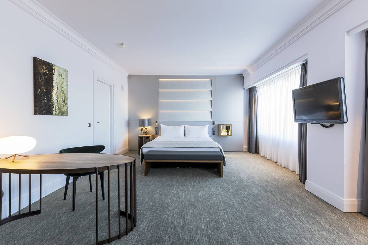 Quarto de hotel moderno com uma cama grande, uma televisão de tela plana, uma mesa de jantar redonda e uma pequena mesa lateral.