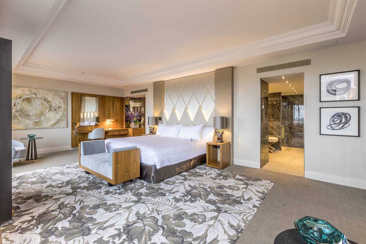 Quarto de hotel moderno com uma cama grande, área de estar com sofá e poltronas, uma pequena mesa e um banheiro visível por uma porta aberta.