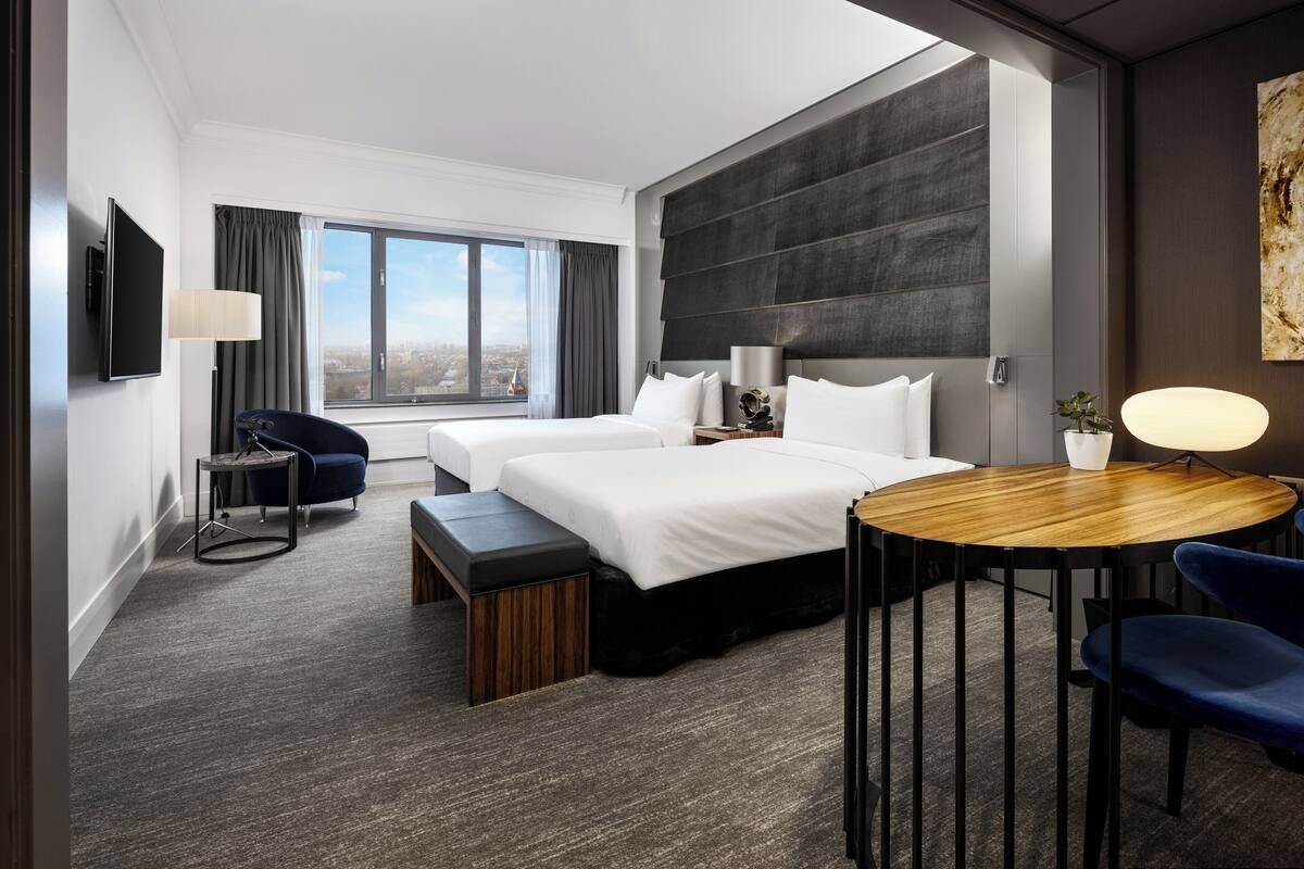 Quarto de hotel com duas camas, uma mesa de jantar, televisão e uma janela grande com cortinas.