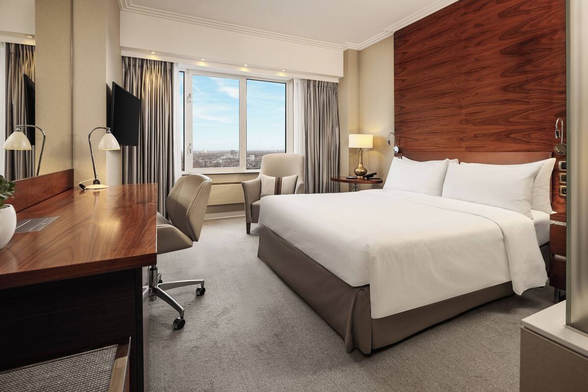 Quarto de hotel com uma cama grande, uma escrivaninha com cadeira, uma televisão e vista para a cidade.