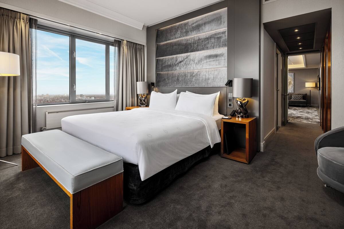 Quarto de hotel moderno com uma cama grande, um banco, vista da cidade e um corredor que dá para outro quarto.