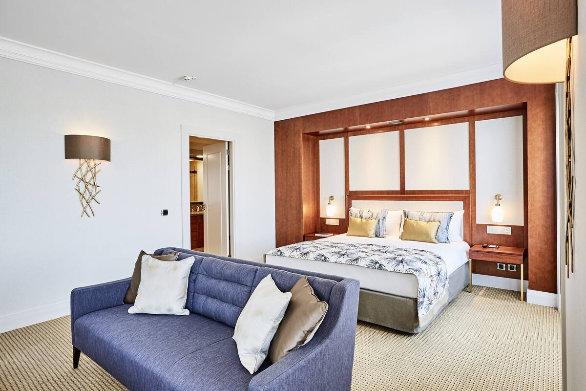 Quarto de hotel com cama, sofá, mesa de cabeceira e luminária fixada na parede.