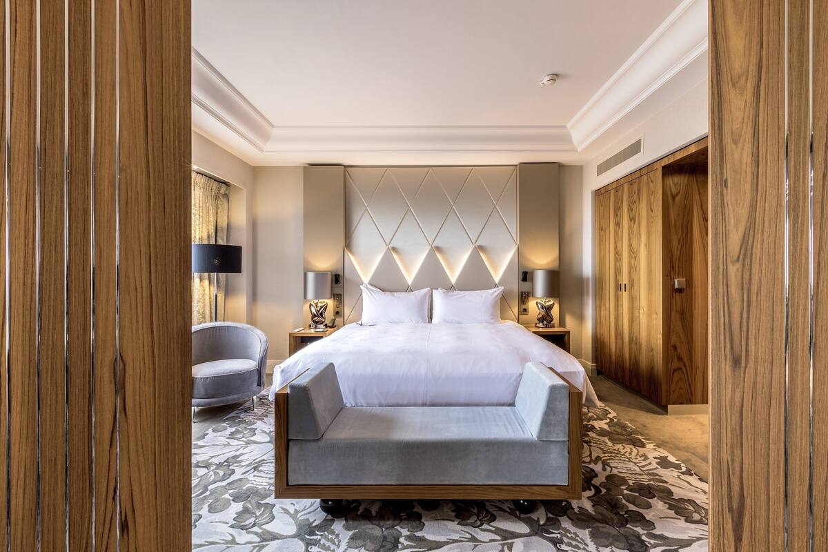 Quarto de hotel moderno com uma cama grande, um sofá cinza, painéis de madeira e um tapete estampado.