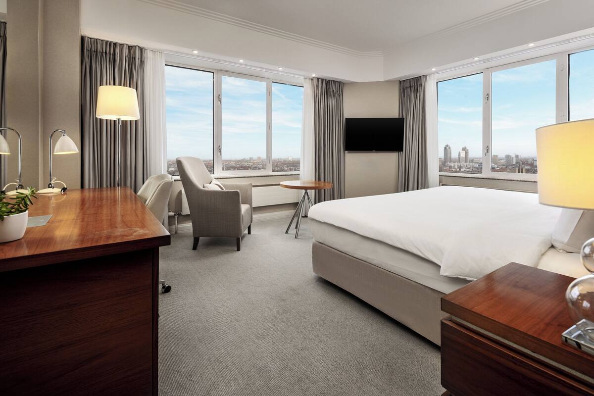 Quarto de hotel com cama grande, escrivaninha com luminária, cadeira, mesa pequena e televisão.