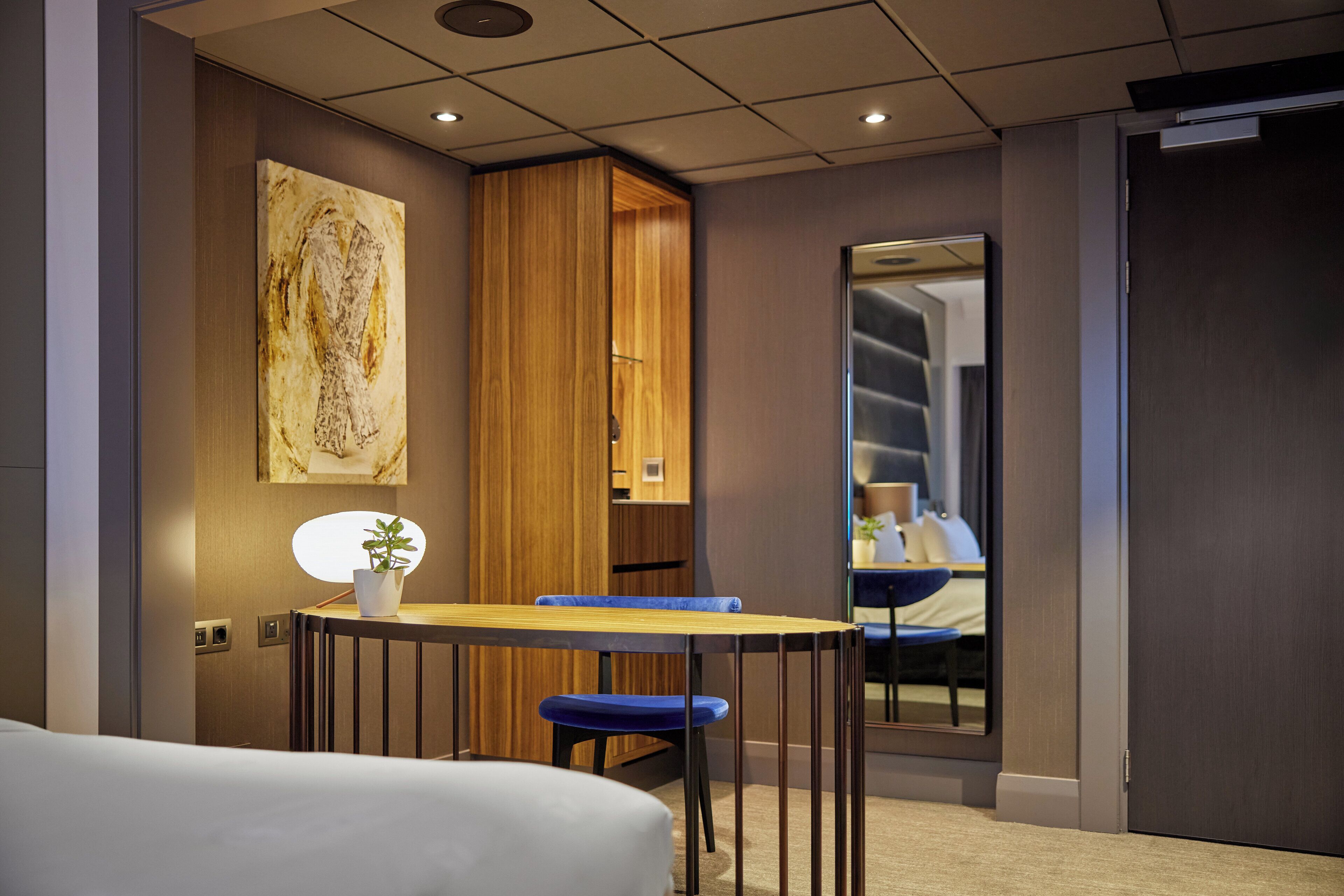 Quarto de hotel moderno com uma escrivaninha de madeira, uma cadeira azul, uma mesa de cabeceira branca e um espelho grande refletindo o quarto.