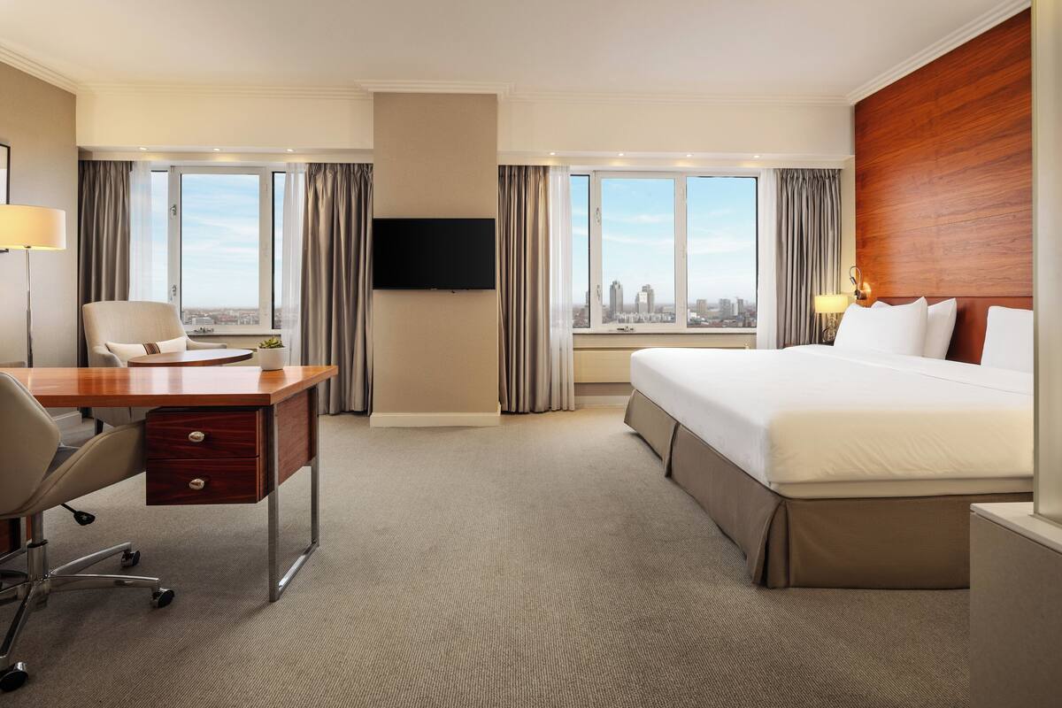 Quarto de hotel com cama, escrivaninha, cadeira e uma janela grande com vista para a cidade.