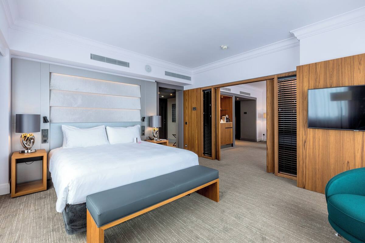 Quarto de hotel moderno com uma cama grande, mesas de cabeceira, um banco, televisão e guarda-roupa de madeira.