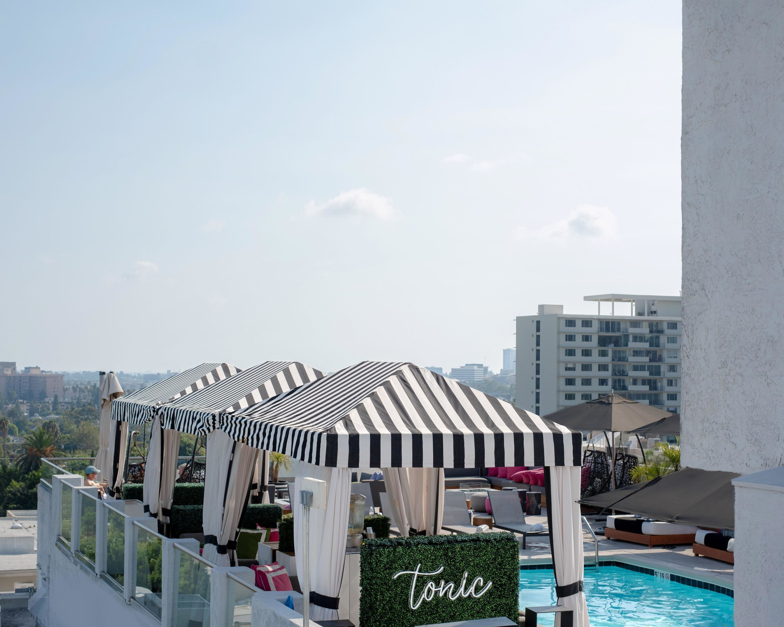 Área de piscina no terraço com toldos listrados, cadeiras de lounge e uma placa com a inscrição 'Tonic'.