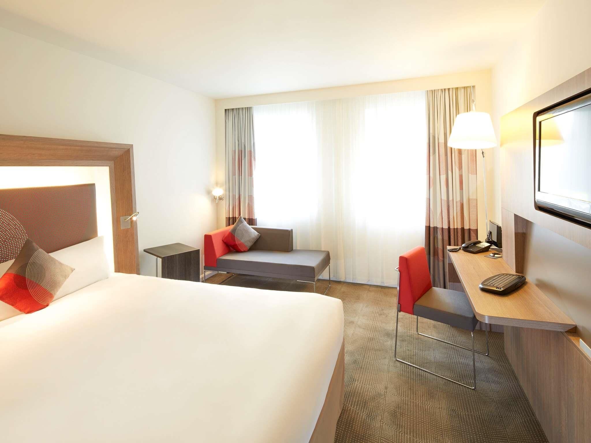 Quarto de hotel com cama, escrivaninha com telefone, cadeira, sofá e televisão.