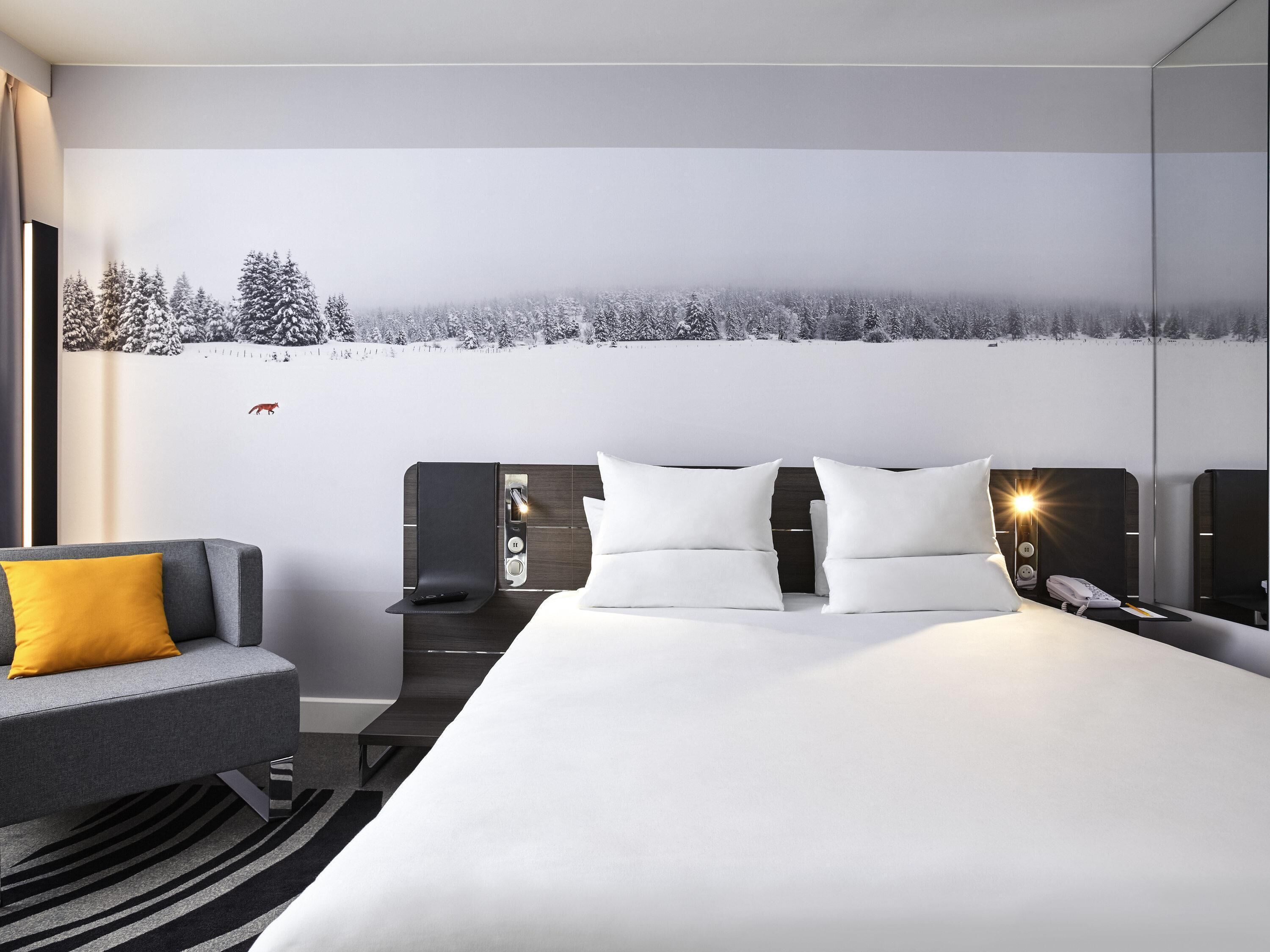 Quarto de hotel moderno com uma cama grande, um sofá cinza com um travesseiro amarelo, uma mesa de cabeceira com uma luminária e um mural na parede retratando uma paisagem nevada.