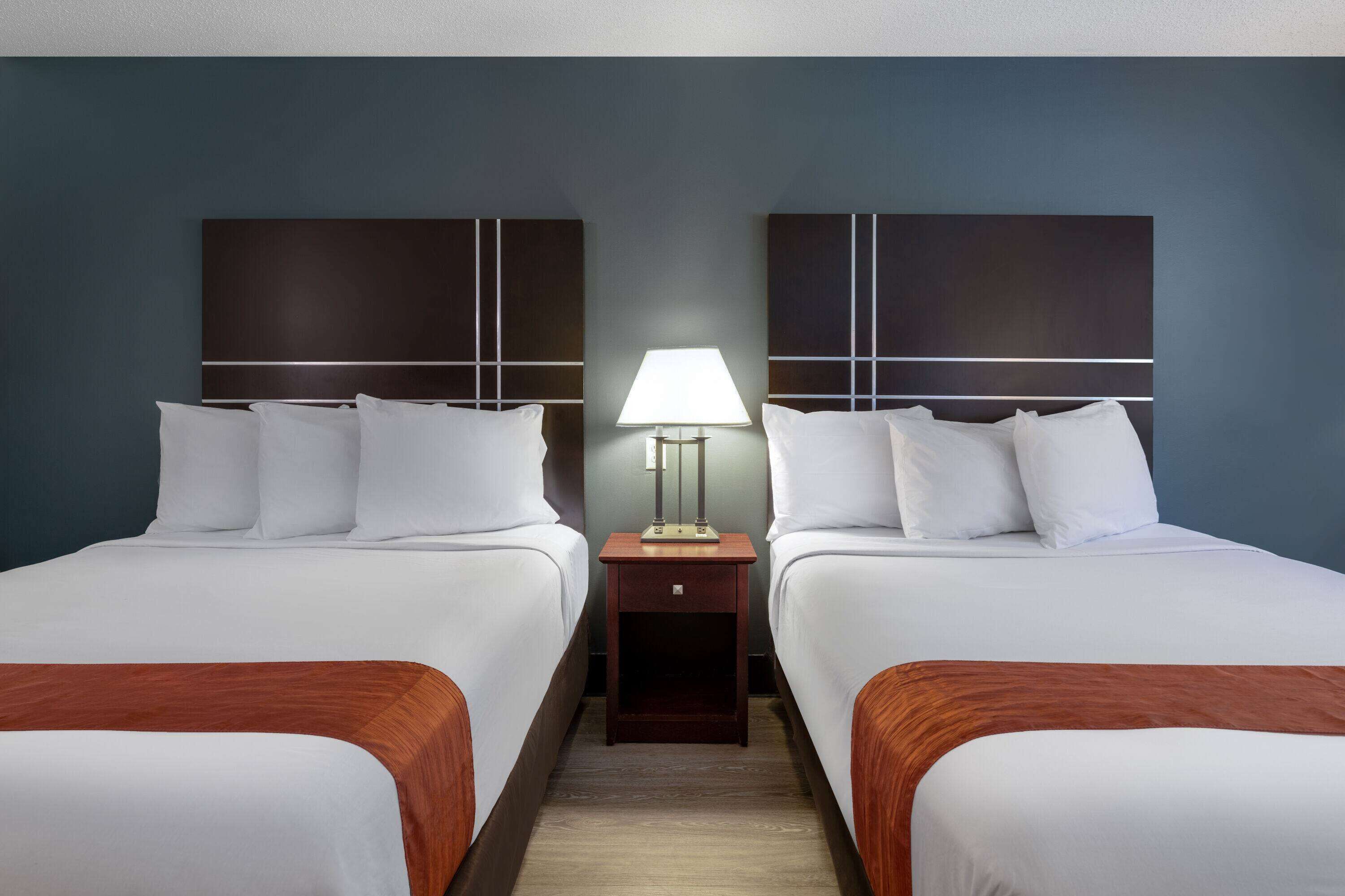 Quarto de hotel com duas camas, mesa de cabeceira com abajur e cabeceira escura.