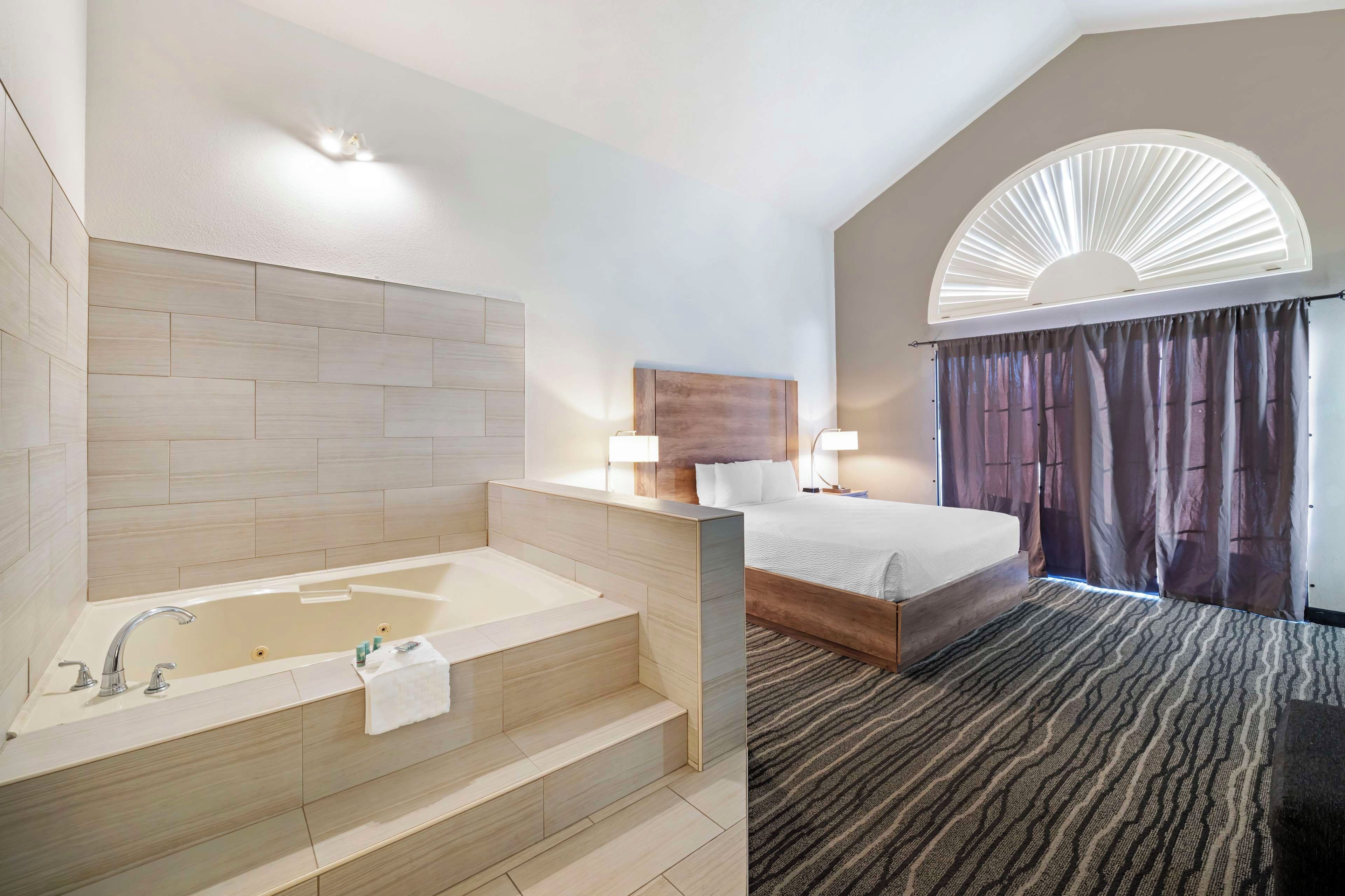 Quarto de hotel com uma banheira grande, uma cama com cabeceira de madeira e vista para o exterior através de uma janela com cortinas.